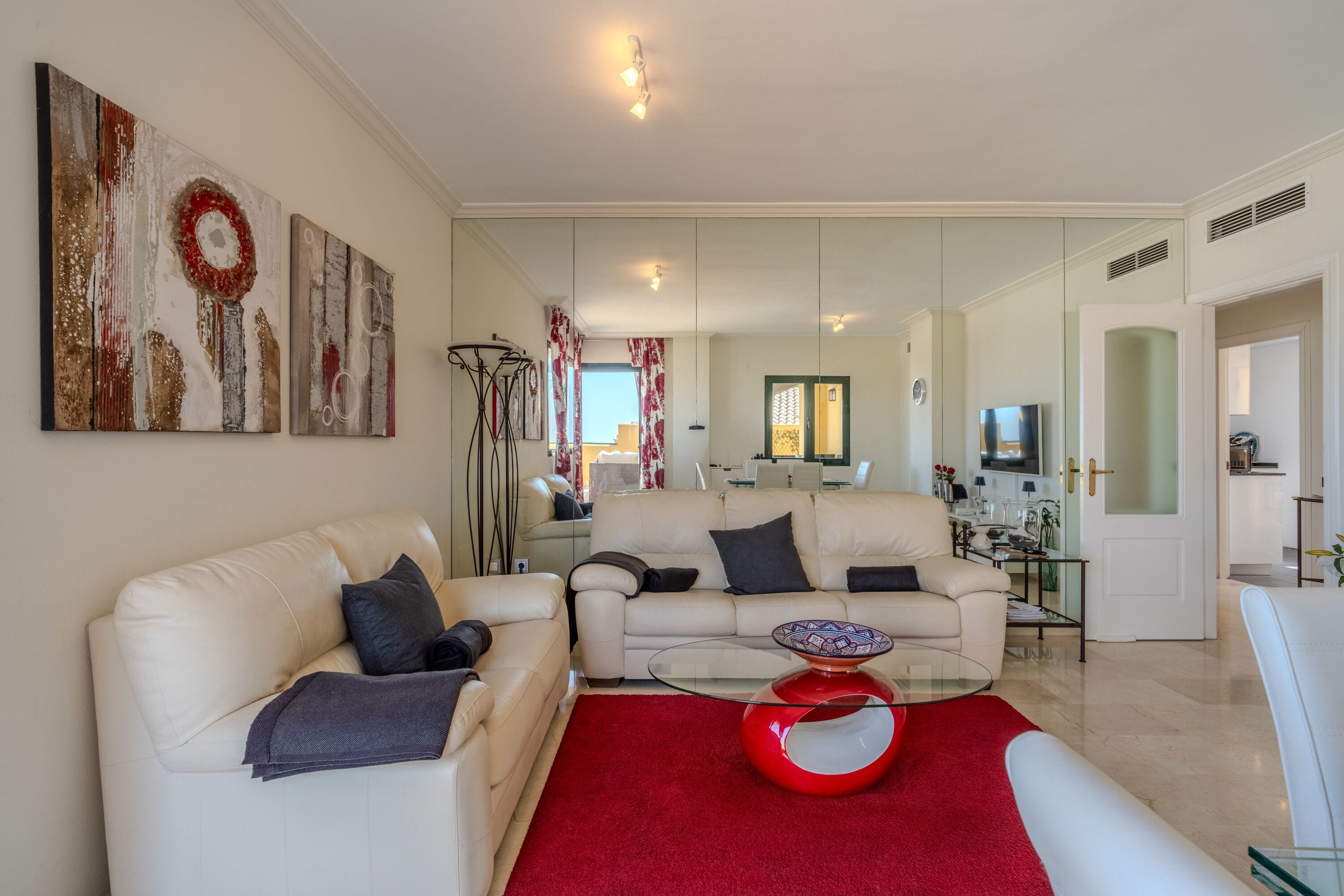 Uma sala de estar moderna com um tapete vermelho, dois sofás brancos, uma mesa de centro de vidro e arte abstrata nas paredes.