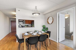 Uma área de refeições com mesa de madeira, cadeiras pretas, luminária pendente, cozinha ao fundo e uma porta branca.