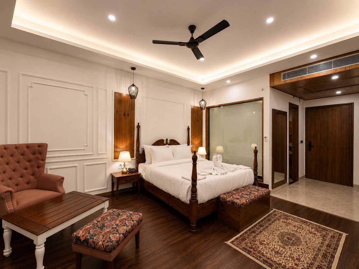 Quarto de hotel com cama, uma cadeira, mesa, banco, ventilador de teto e um espelho grande.