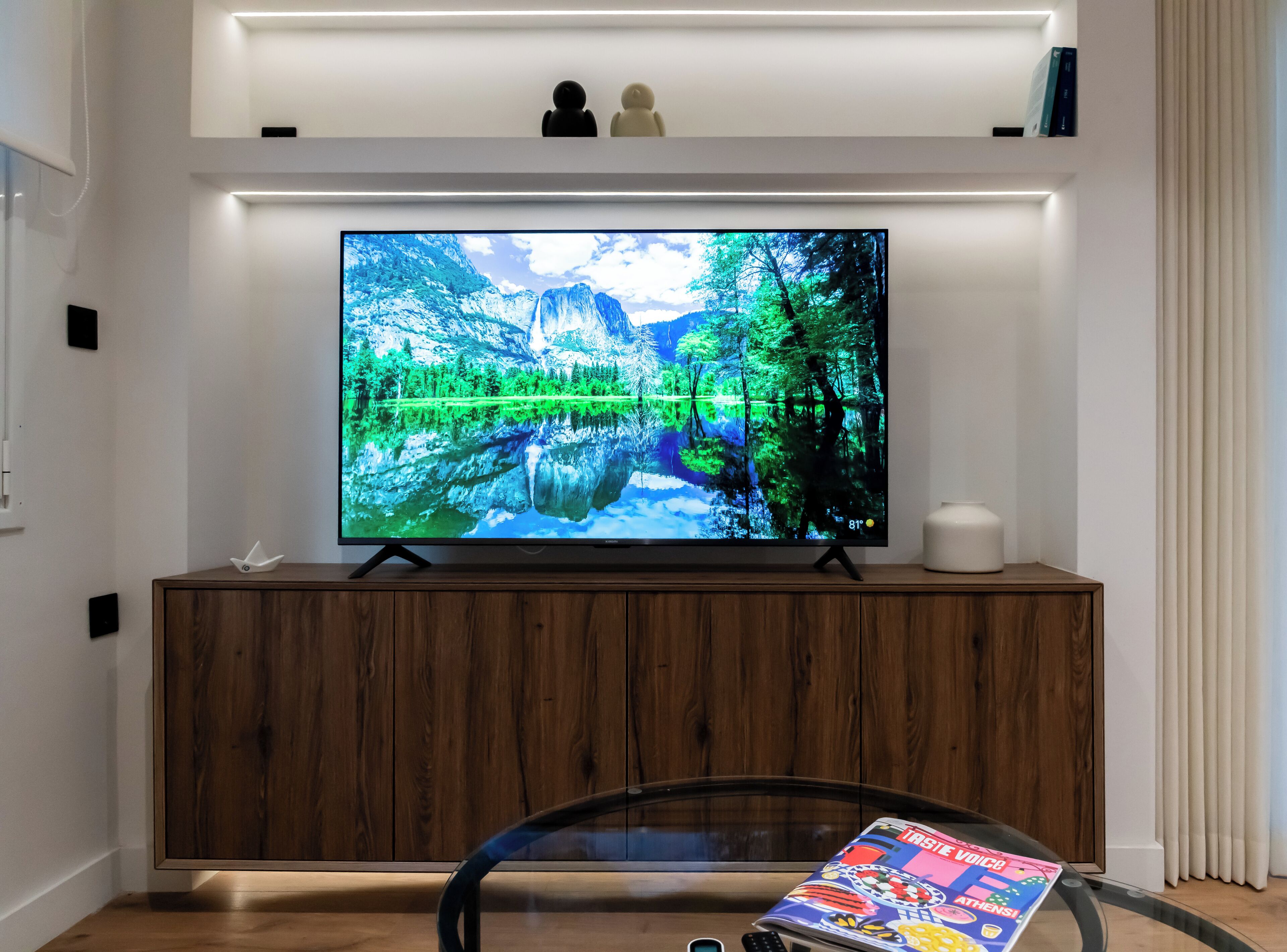 Uma TV de tela plana fixada em um armário de madeira, com uma revista sobre uma mesa de vidro à frente.