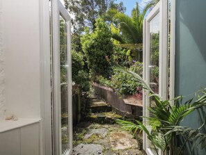 Um caminho estreito no jardim leva a uma porta, cercado por vegetação exuberante e um pequeno elemento aquático.