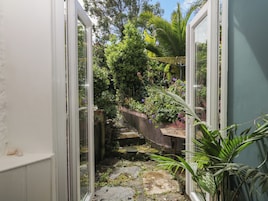 Um caminho estreito no jardim leva a uma porta, cercado por vegetação exuberante e um pequeno elemento aquático.