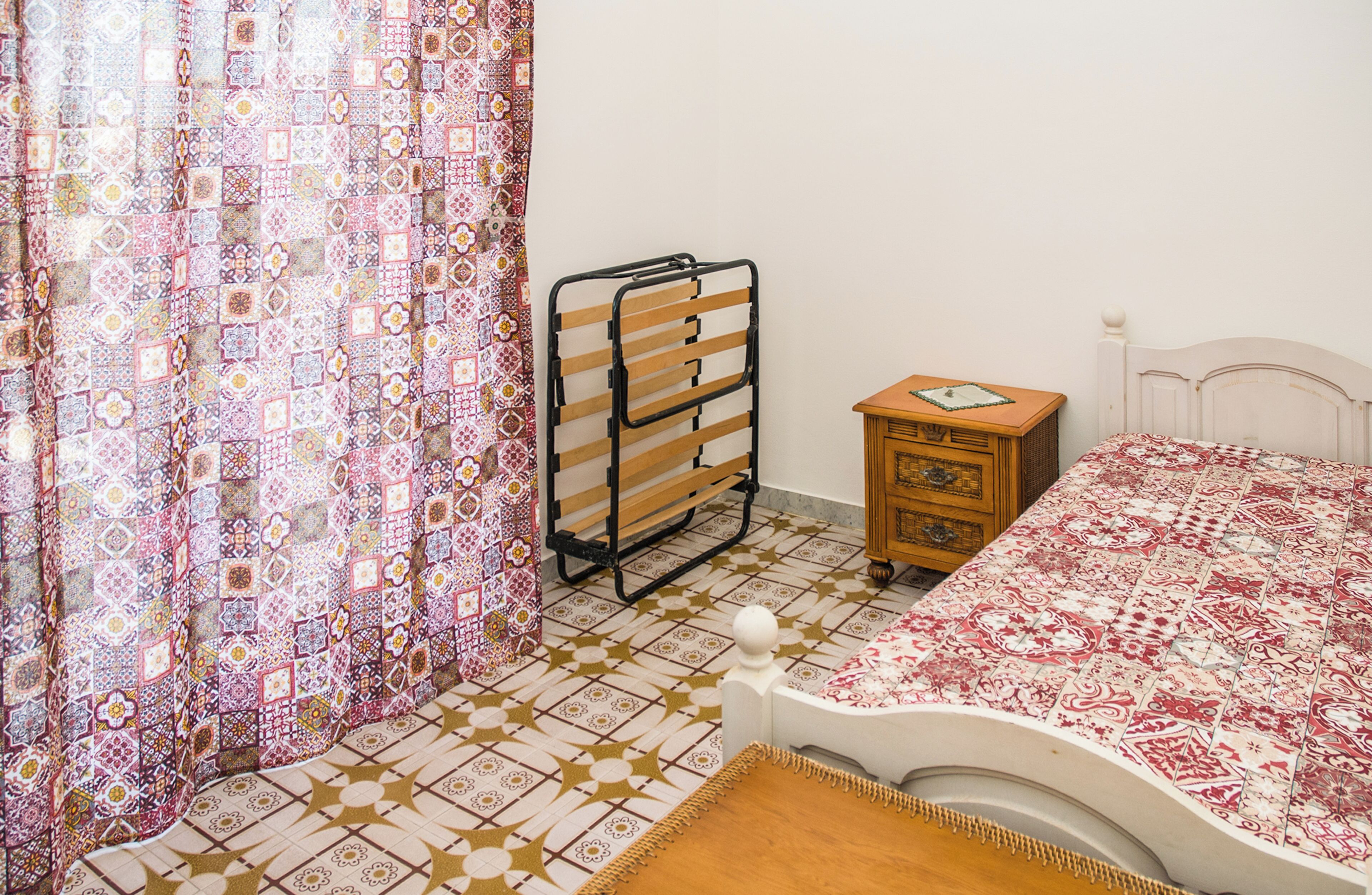 Quarto com colcha estampada, mesa de cabeceira de madeira, varal de metal e cortina padronada.
