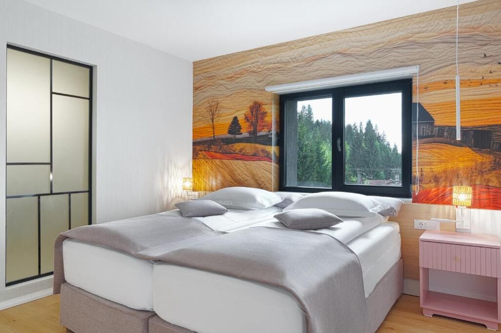 Quarto com cama, janela e parede com painel de paisagem.