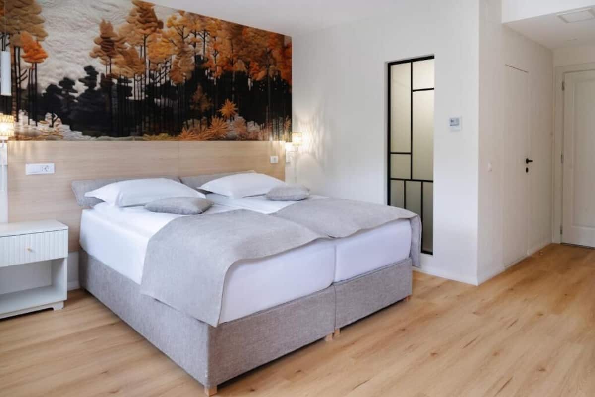 Um quarto com cama, mesa de cabeceira, luminária na parede e uma porta.