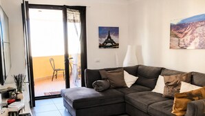 Uma sala de estar com um sofá seccional cinza, uma mesinha com abajur e uma porta que dá para uma varanda.