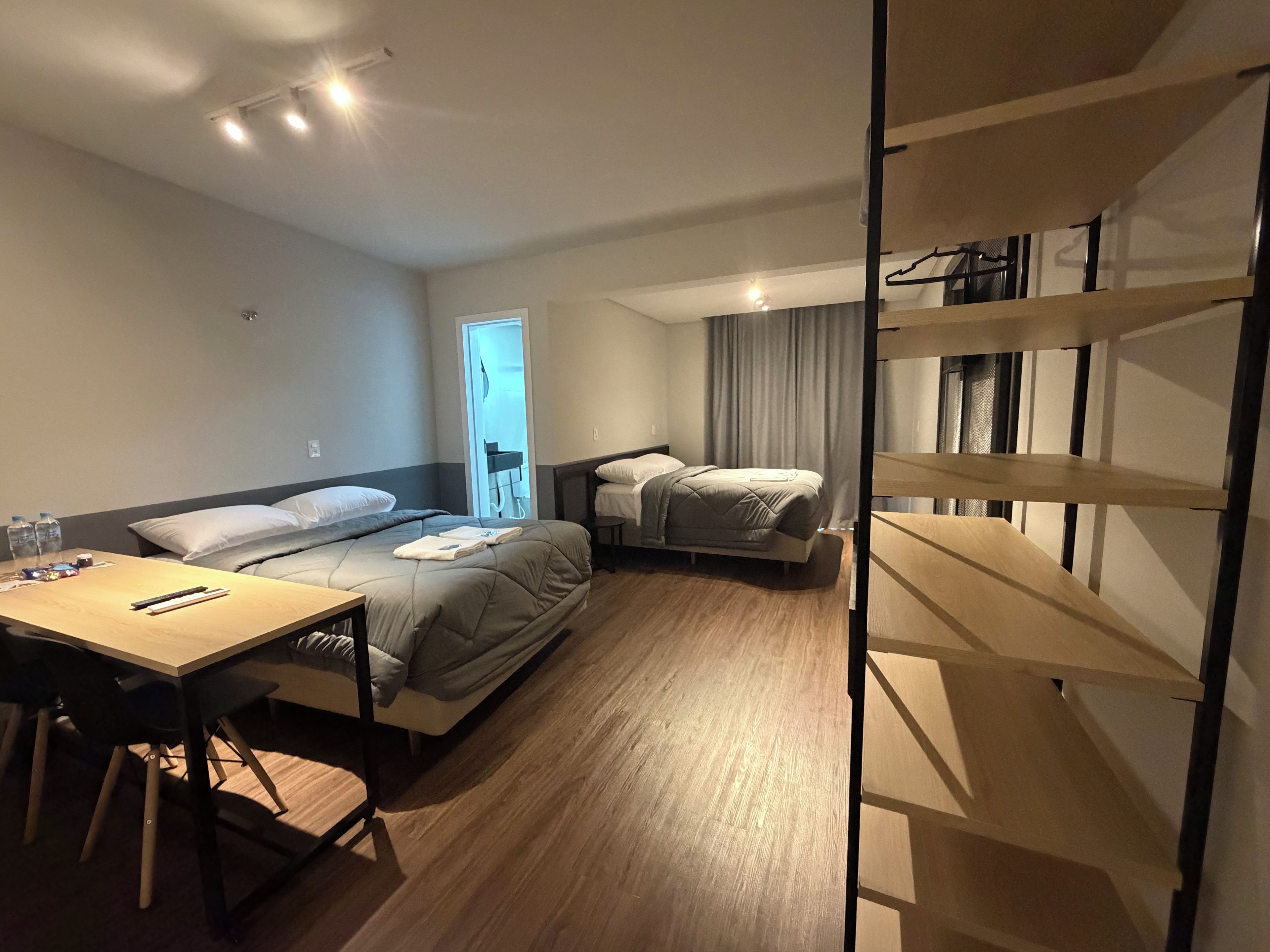 Quarto de hotel com cama, escrivaninha e cadeira. Há uma janela com cortinas e uma porta que dá para outro quarto.