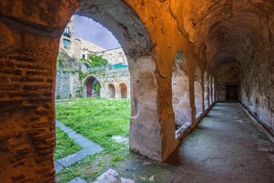 Um corredor com arcos, um pátio ajardinado e um edifício ao fundo.