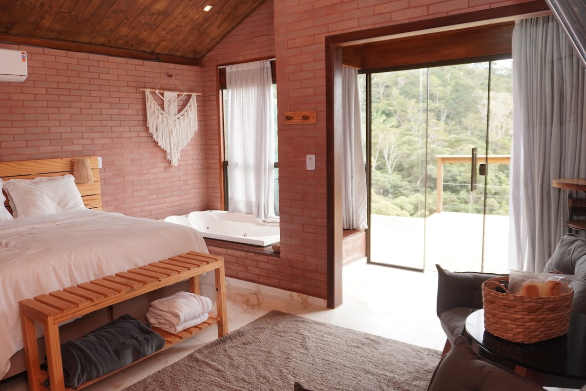 Quarto com cama de madeira, um banco, janela com cortinas e vista para vegetação lá fora.