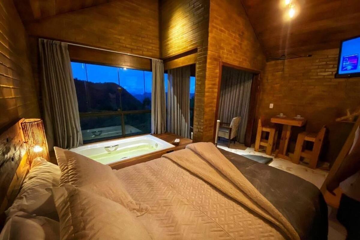 Quarto com cama grande, janela com cortinas, mesa pequena e televisão na parede.