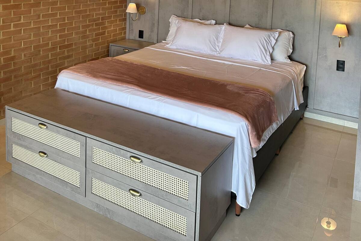 Quarto com parede de tijolos, teto de madeira, cama com cabeceira cinza, abajures e um móvel embutido.