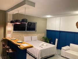 Quarto de hotel com uma cama, uma escrivaninha com cadeiras, uma luminária e uma janela com grade de metal.