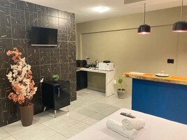 Quarto de hotel moderno com um armário preto, uma escrivaninha com micro-ondas, uma televisão na parede, um balcão de recepção e uma planta em vaso.