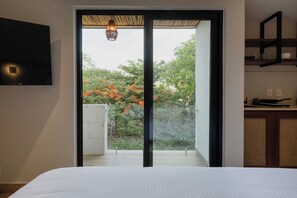 Quarto com cama, televisão na parede e porta deslizante para uma varanda com vista para árvores e flores.