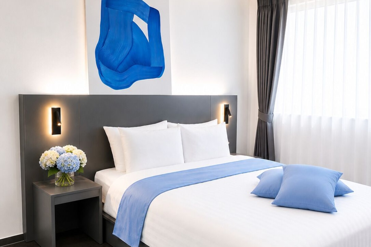 Quarto de hotel moderno com uma cama grande, uma mesa de cabeceira com um vaso de flores e uma pintura abstrata azul na parede.