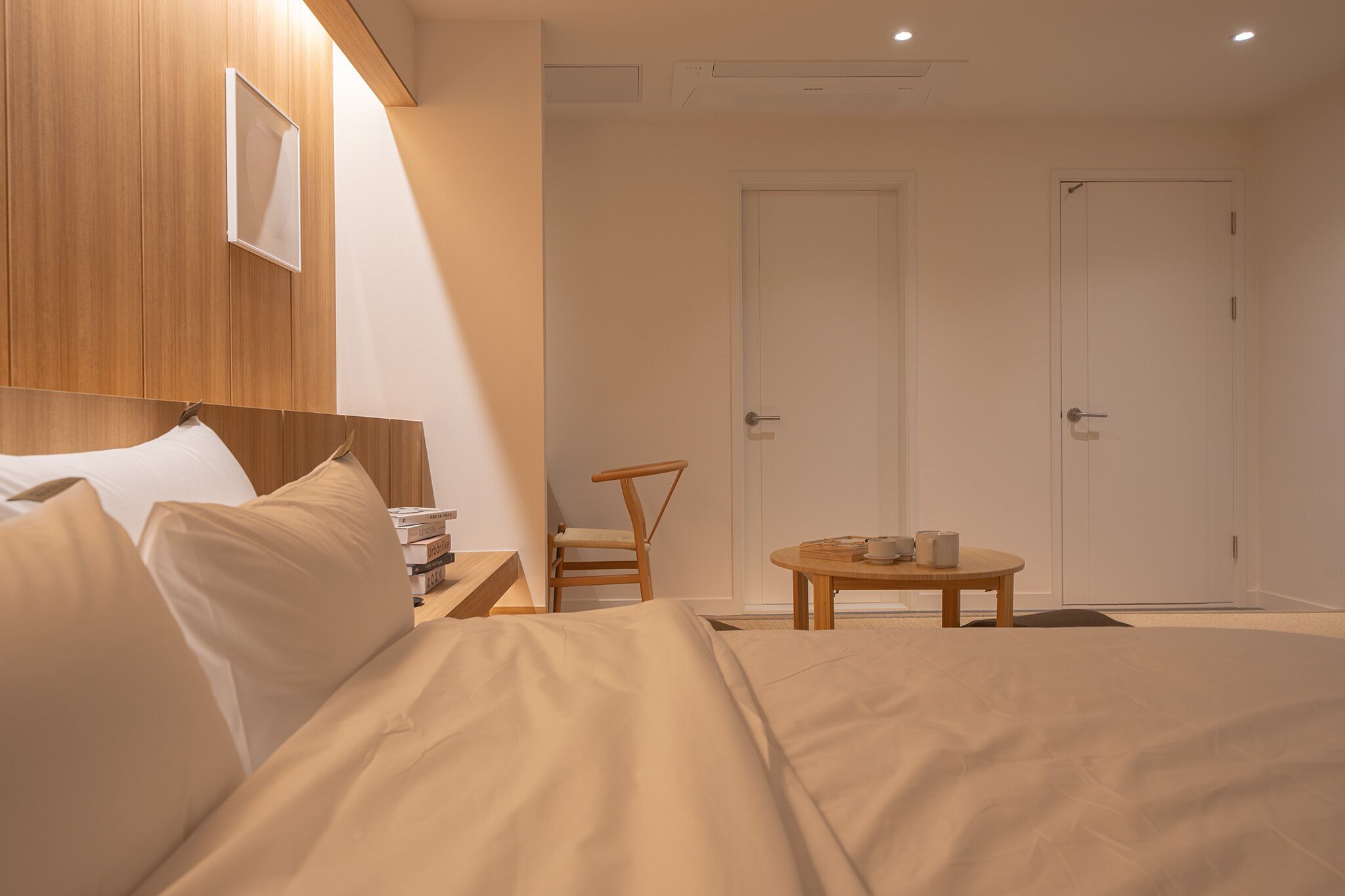 Quarto de hotel com cama, cabeceira de madeira, uma cadeira, uma mesinha e duas portas.