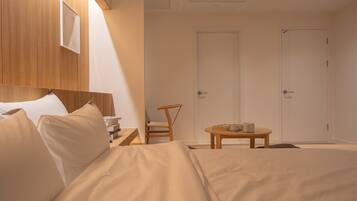 Quarto de hotel com cama, cabeceira de madeira, uma cadeira, uma mesinha e duas portas.