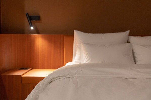 Cama bem arrumada com roupa de cama branca, mesa de cabeceira de madeira e luminária fixada na parede.