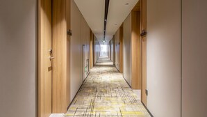 Um corredor de hotel comprido e estreito, com portas de madeira em ambos os lados, carpete estampado e teto com iluminação embutida.