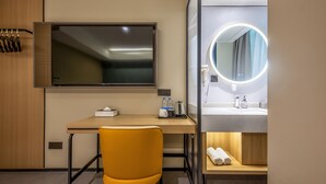 Quarto de hotel com escrivaninha de madeira, uma cadeira amarela, televisão de tela plana, espelho redondo com iluminação embutida, pia e porta-toalhas.