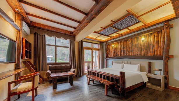 Quarto com cama de madeira, um banco de madeira, sofá, televisão e janelas amplas.