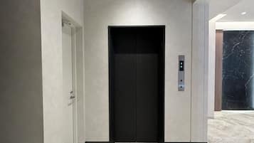 Um elevador em um corredor com piso de mármore, uma porta à esquerda e uma parede com padrão de mármore à direita.