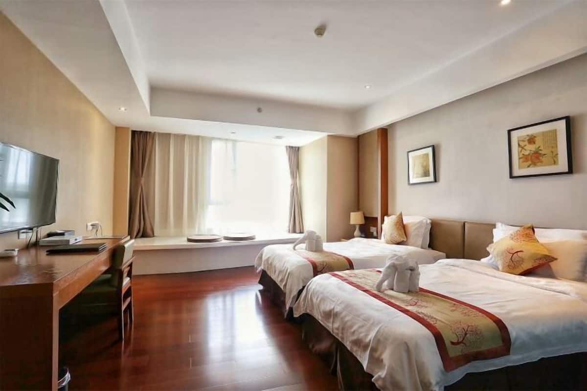 Quarto de hotel com duas camas, uma escrivaninha, televisão e janela com cortinas.