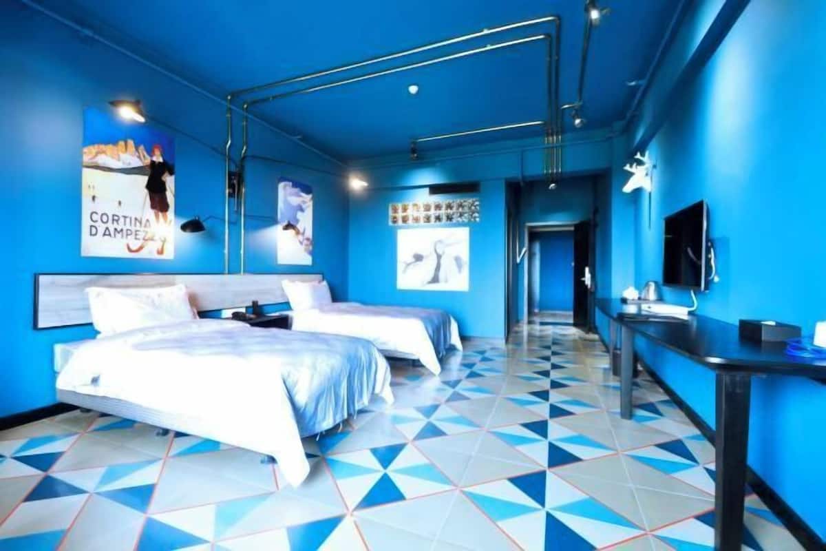 Quarto de hotel com duas camas, uma escrivaninha, televisão e banheiro ao fundo.