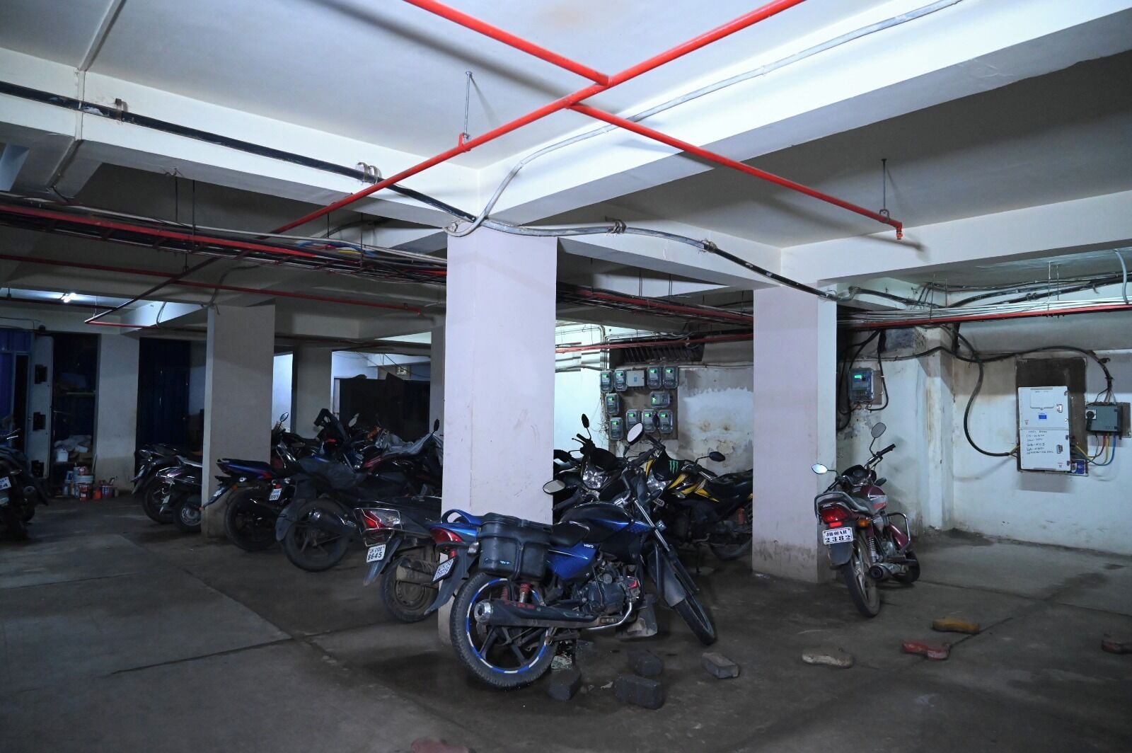 Uma garagem de estacionamento com várias motocicletas estacionadas sob pilares de concreto, fiação aparente e teto com canos vermelhos.