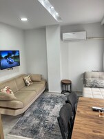 Uma sala de estar moderna com um sofá bege, um tapete estampado, uma mesa de jantar de madeira e uma televisão fixada na parede.