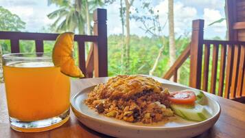 Uma mesa com um prato de arroz frito e um copo de bebida de laranja, com vista para áreas verdes e um corrimão de madeira.