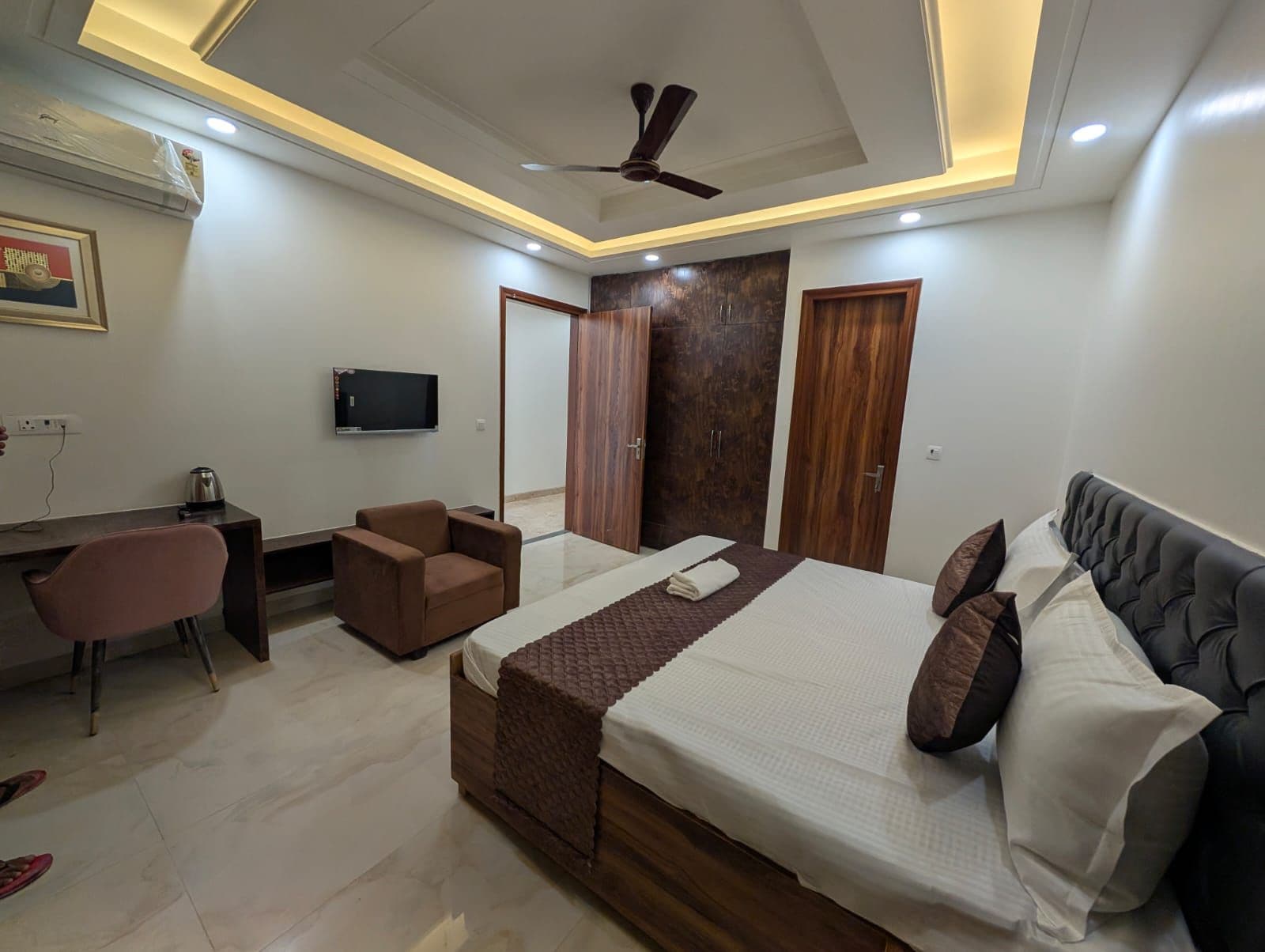 Quarto de hotel com cama, escrivaninha, cadeira, televisão e ventilador de teto.