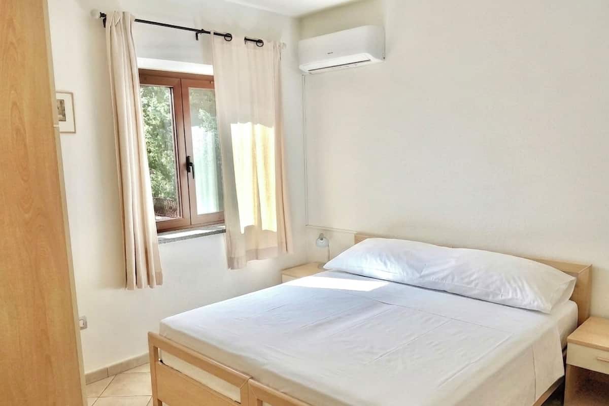 Um quarto com cama, janela com cortinas, mesa de cabeceira e aparelho de ar condicionado.