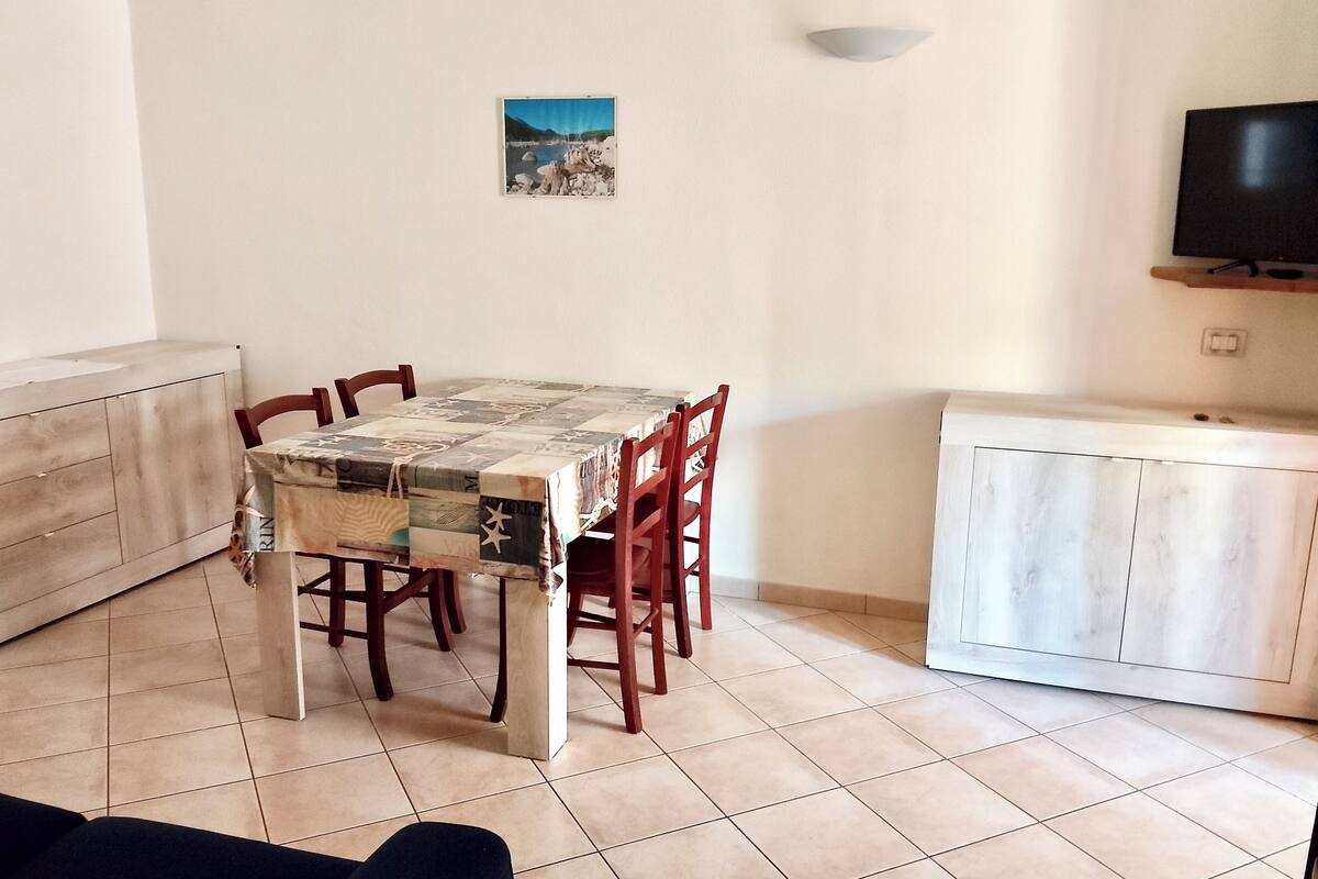 Uma área de refeições com uma mesa coberta por um manto de retalhos, cadeiras de madeira, uma pequena luminária na parede e uma televisão sobre um suporte.