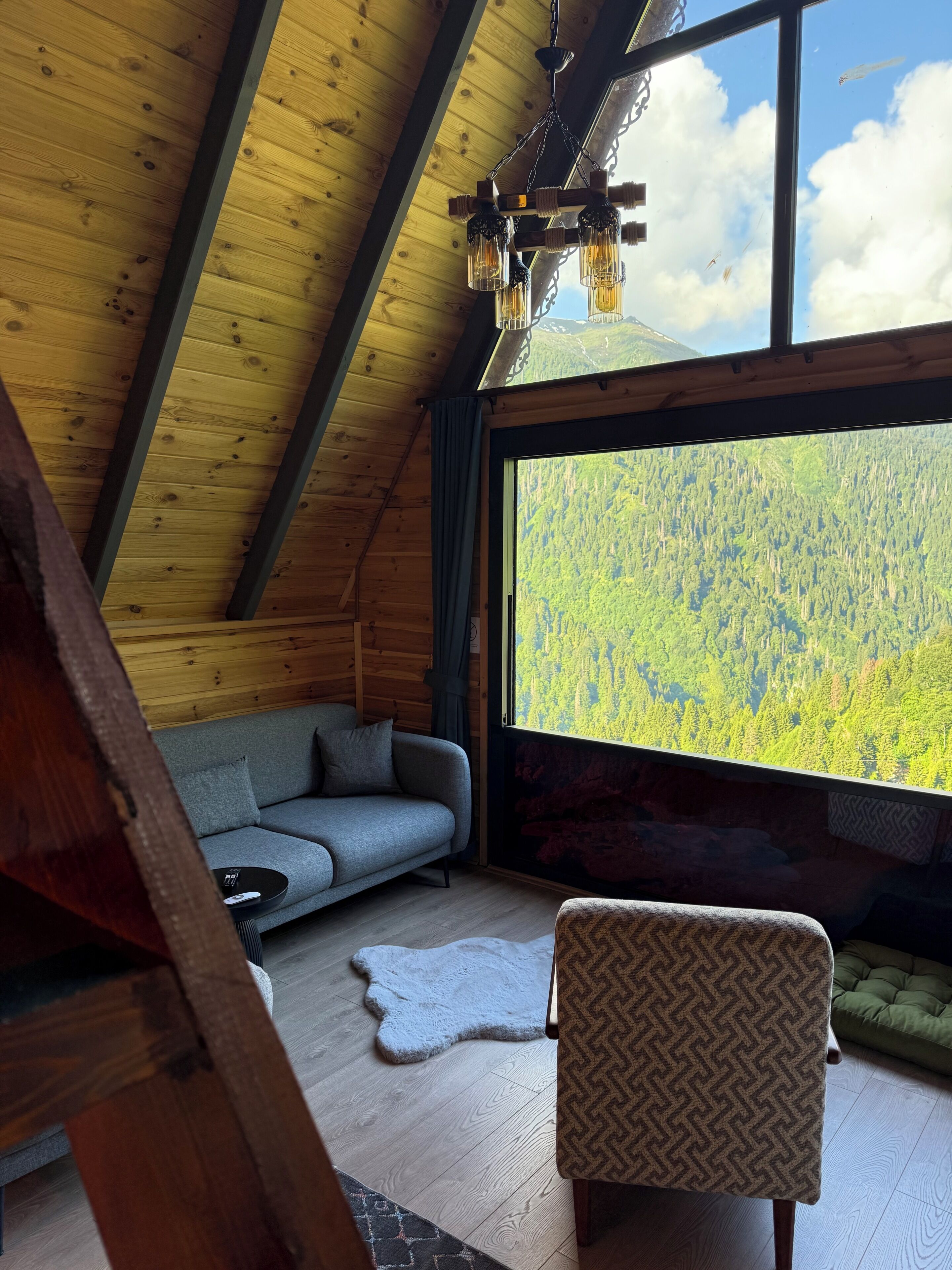 Interior acolhedor de cabana com teto de madeira, um sofá, uma poltrona estampada e uma janela grande com vista para uma floresta.