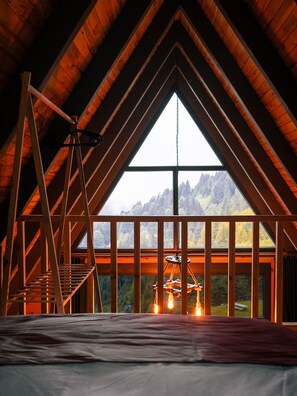 Um quarto com pé direito alto, uma grande janela com vista para uma paisagem montanhosa, uma escada de madeira e um lustre.