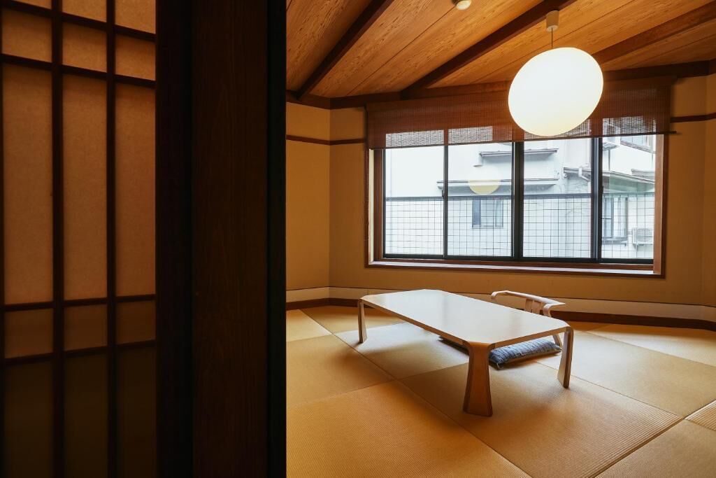 A traditional Japanese room with tatami flooring, a low wooden table, and a large window with a view of buildings.