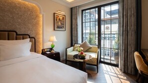 Quarto de hotel com cama, sofá, mesinha de centro com abajur, quadro emoldurado e janela com cortinas.