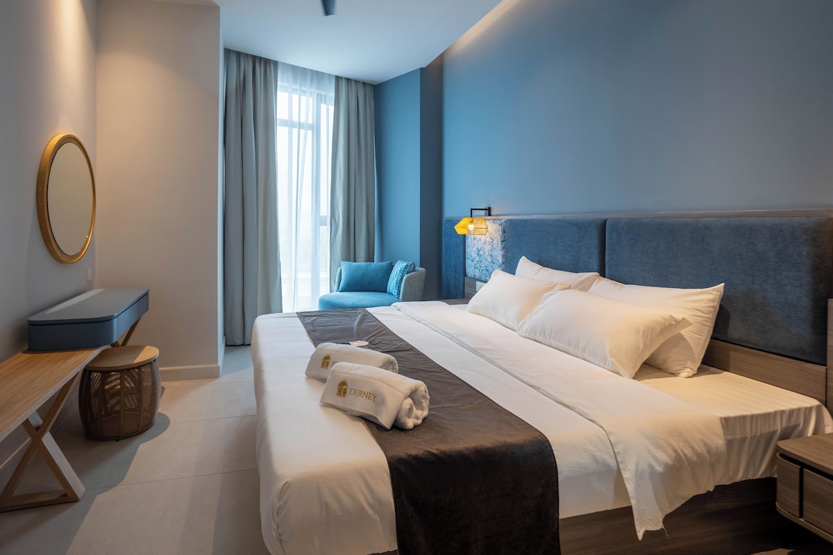 Quarto de hotel com uma cama grande, uma mesinha de cabeceira, uma cadeira, uma janela com cortinas e uma luminária na parede.