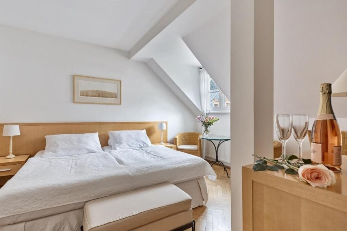 Quarto de hotel com cama, mesa de cabeceira, cadeira, pequena mesa com flores e uma garrafa de champanhe sobre um armário de madeira.