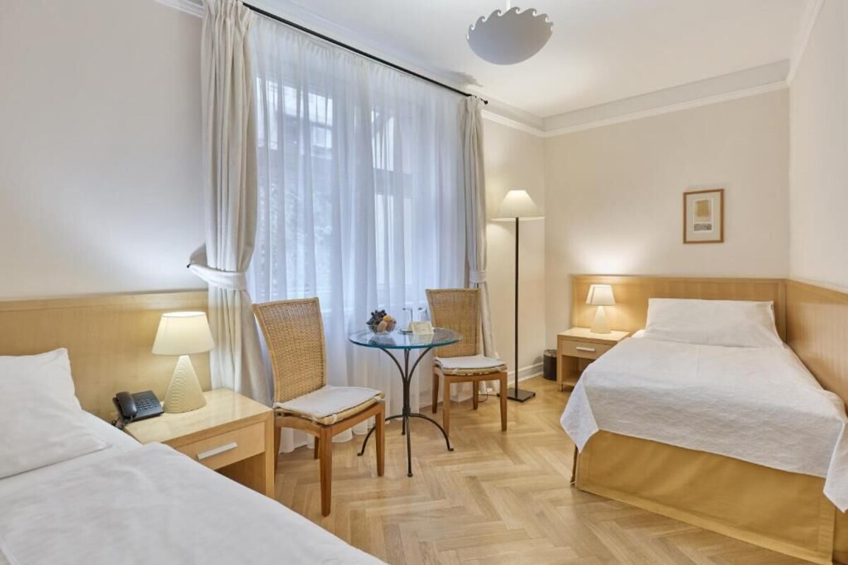 Quarto de hotel com duas camas, uma mesa pequena com cadeiras, um abajur, mesa de cabeceira com telefone e um quadro na parede.