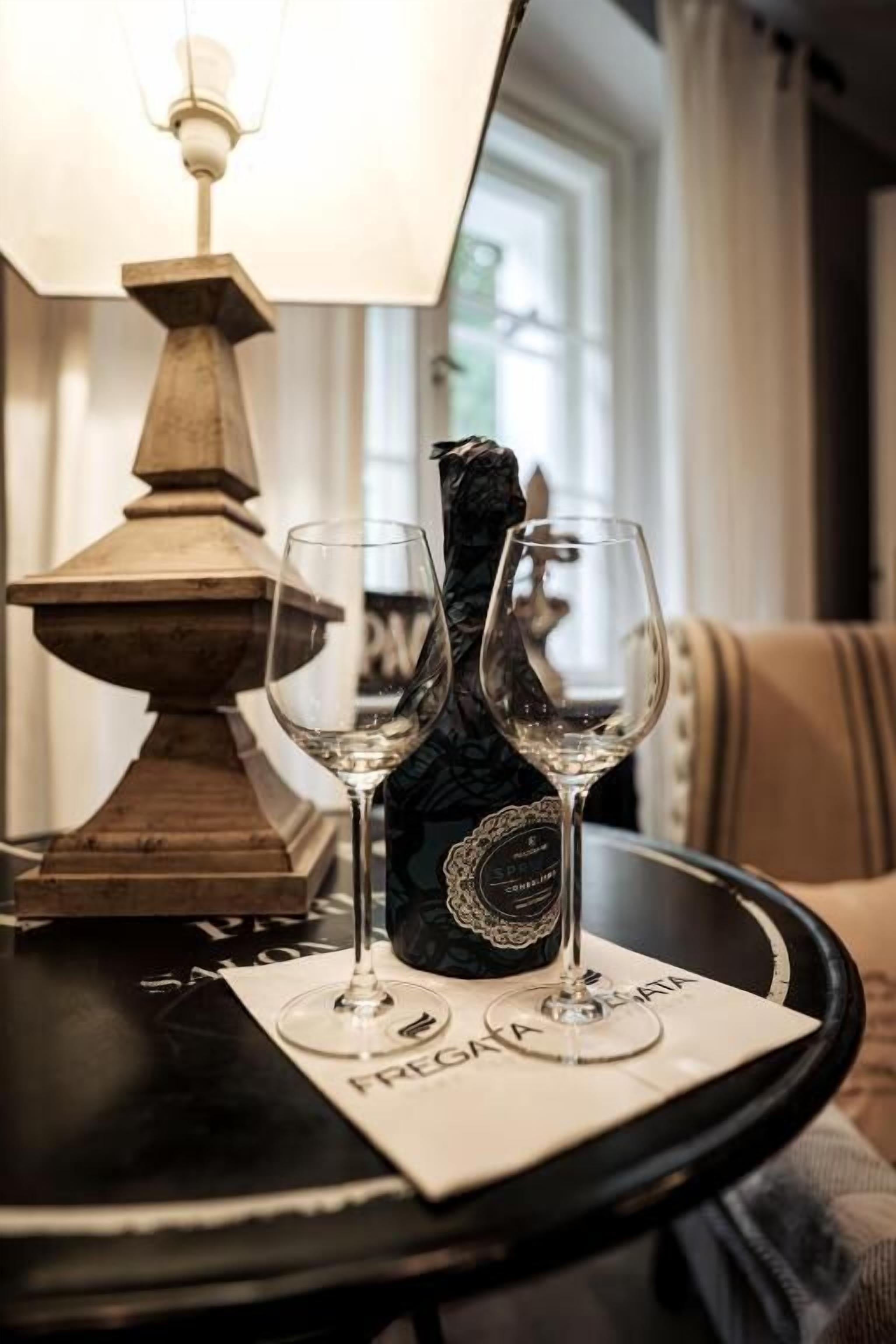 Uma mesa com duas taças de vinho e uma garrafa, um abajur e uma cadeira ao fundo.