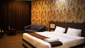 Quarto de hotel com uma cama grande, uma poltrona azul escura, uma pequena mesa redonda e um papel de parede estampado.