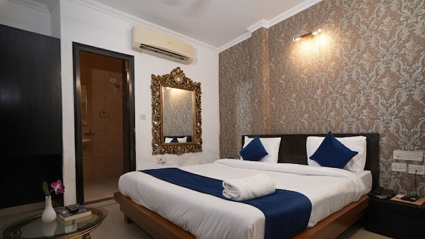 Quarto de hotel com uma cama grande, uma mesa de cabeceira com um vaso e um livro, um espelho e papel de parede estampado.