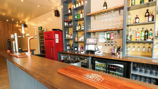 Uma área de bar com balcão de madeira, prateleiras com garrafas e um refrigerador.