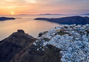Uma vila costeira com edifícios brancos, uma colina rochosa e um mar calmo ao pôr do sol.