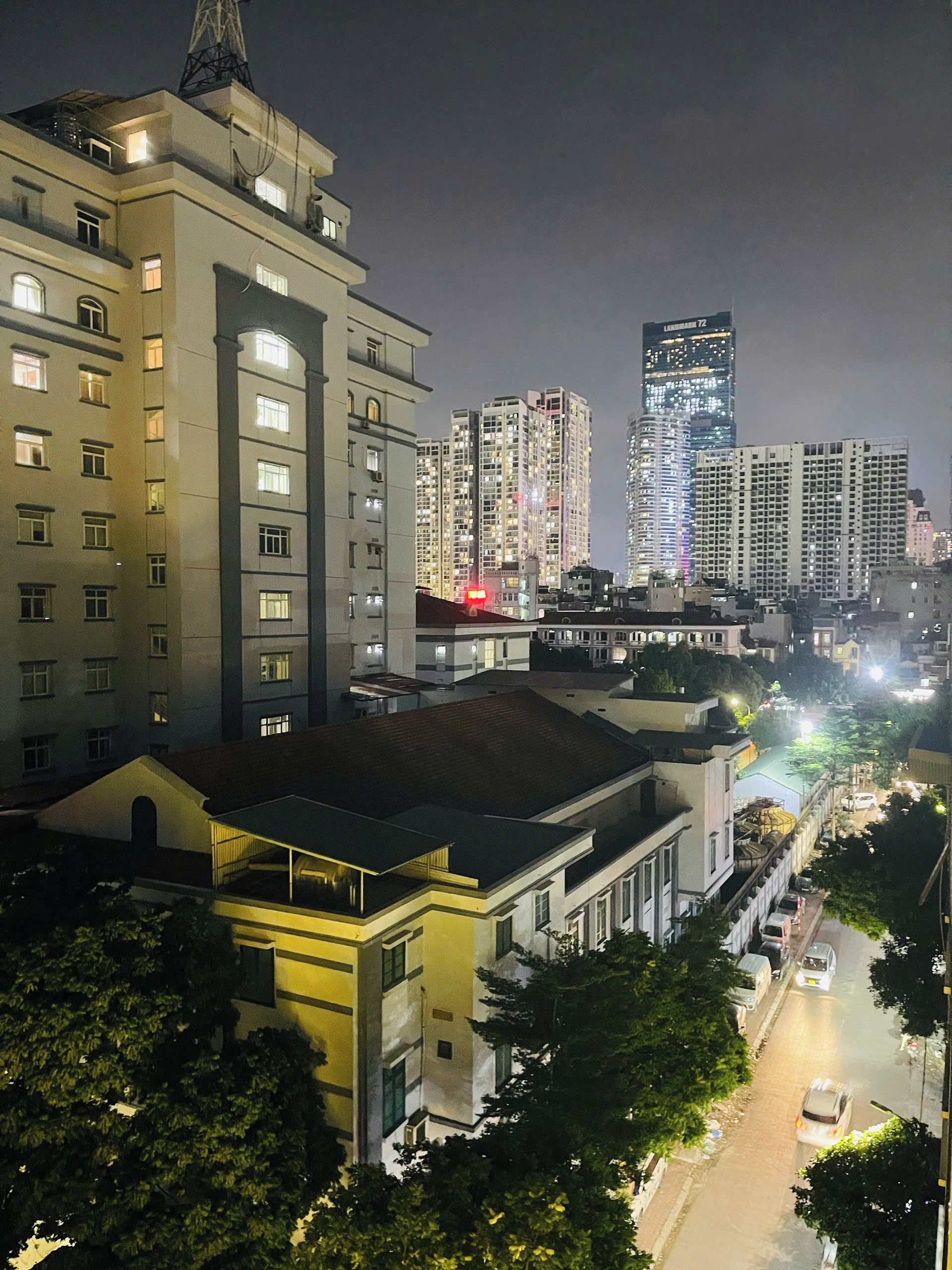 Paisagem urbana noturna com arranha-céus, uma torre imponente e uma rua com carros estacionados.