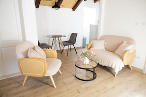 Uma sala de estar acolhedora, com um sofá, duas poltronas, uma mesinha redonda com um vaso, e um espaço de jantar com mesa e cadeiras.
