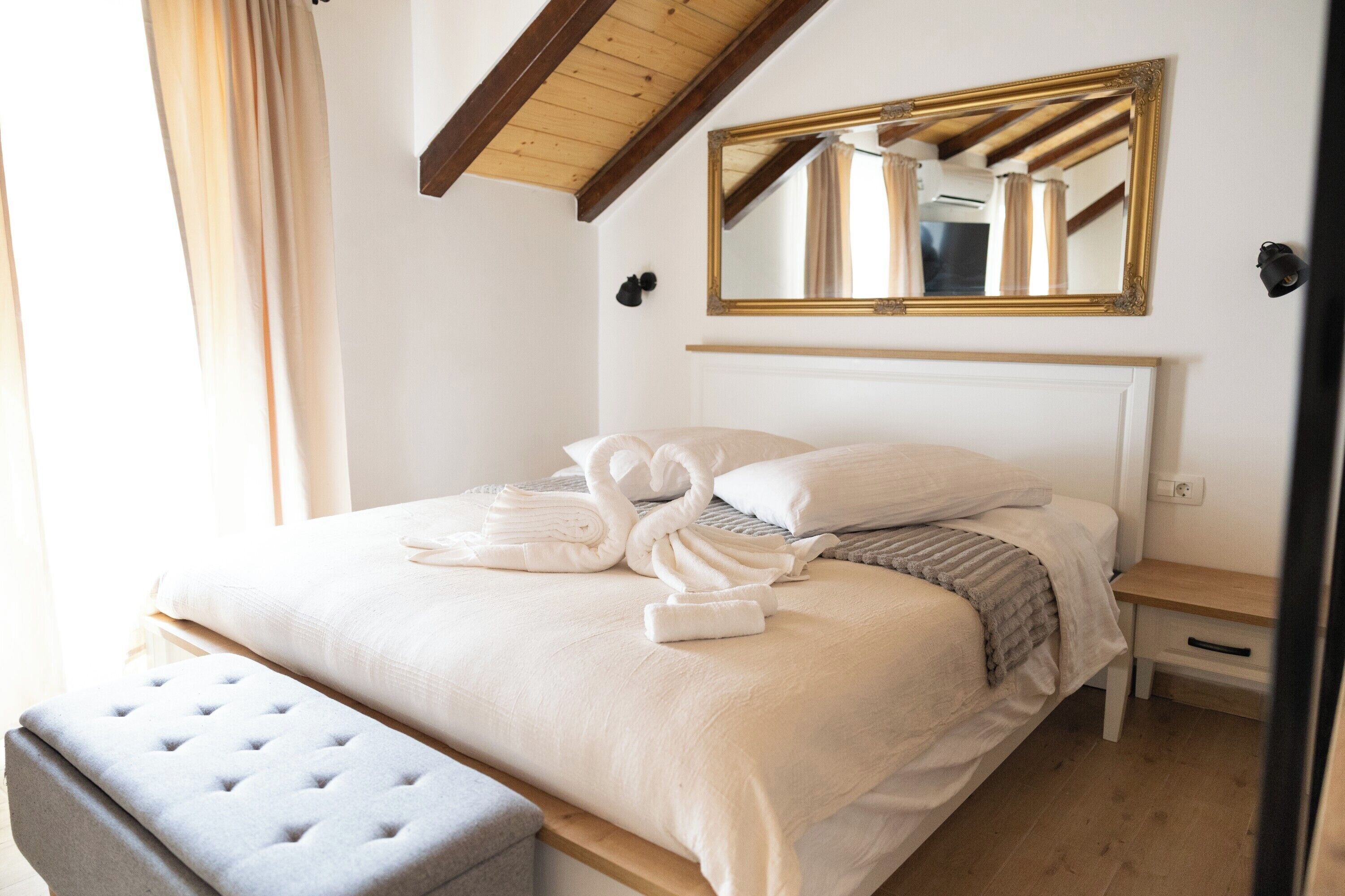 Quarto de hotel com cama, mesa de cabeceira, criado-mudo, banquinho e espelho.