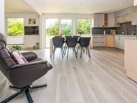 Uma sala de estar moderna com sofá, mesa de jantar e cadeiras, cozinha com armários e eletrodomésticos, e grandes janelas com vista para árvores.